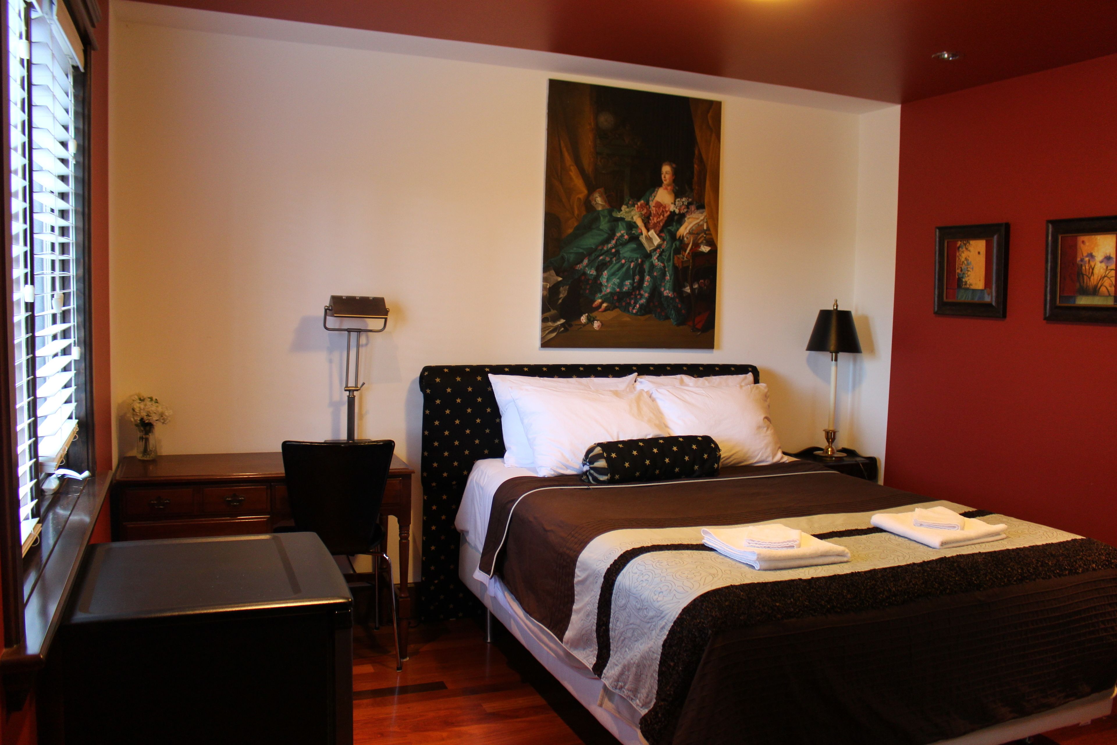 Quarto de hotel com cama, escrivaninha, cadeira, abajur e um quadro na parede.