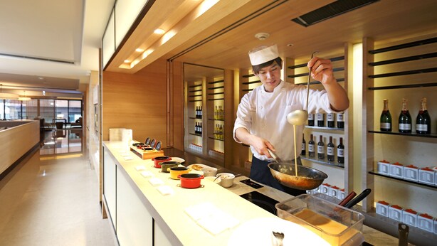 Um chef cozinha em uma cozinha moderna com detalhes em madeira e um balcão comprido exibindo diversos utensílios e ingredientes culinários.