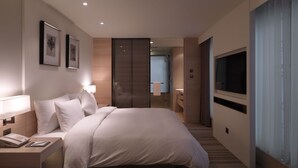 Quarto de hotel moderno com uma cama grande, mesas de cabeceira, televisão e área de chuveiro.