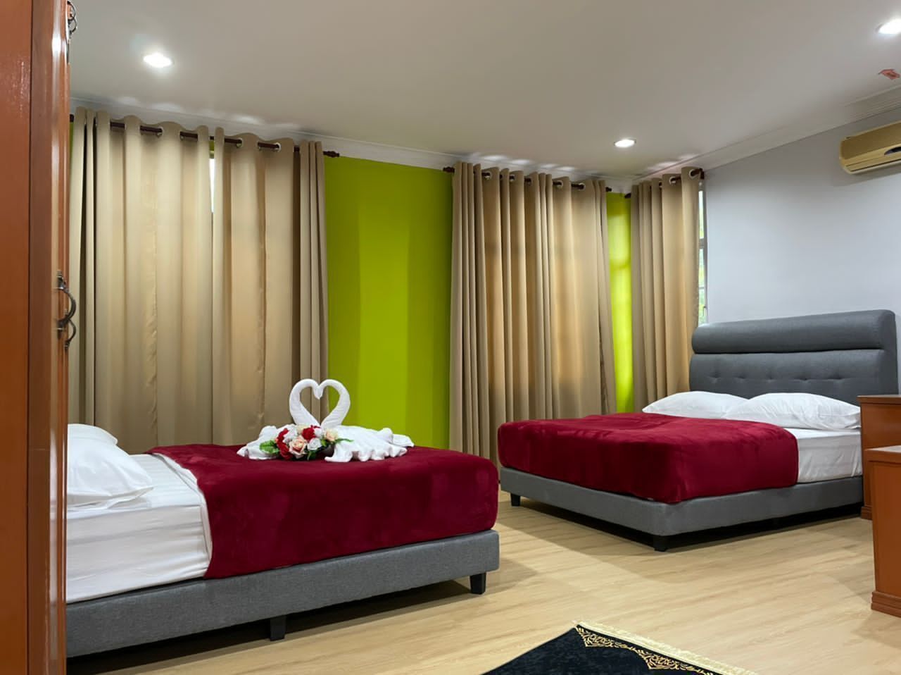 Quarto de hotel com duas camas, cortinas bege, uma parede de destaque verde e uma cabeceira cinza.
