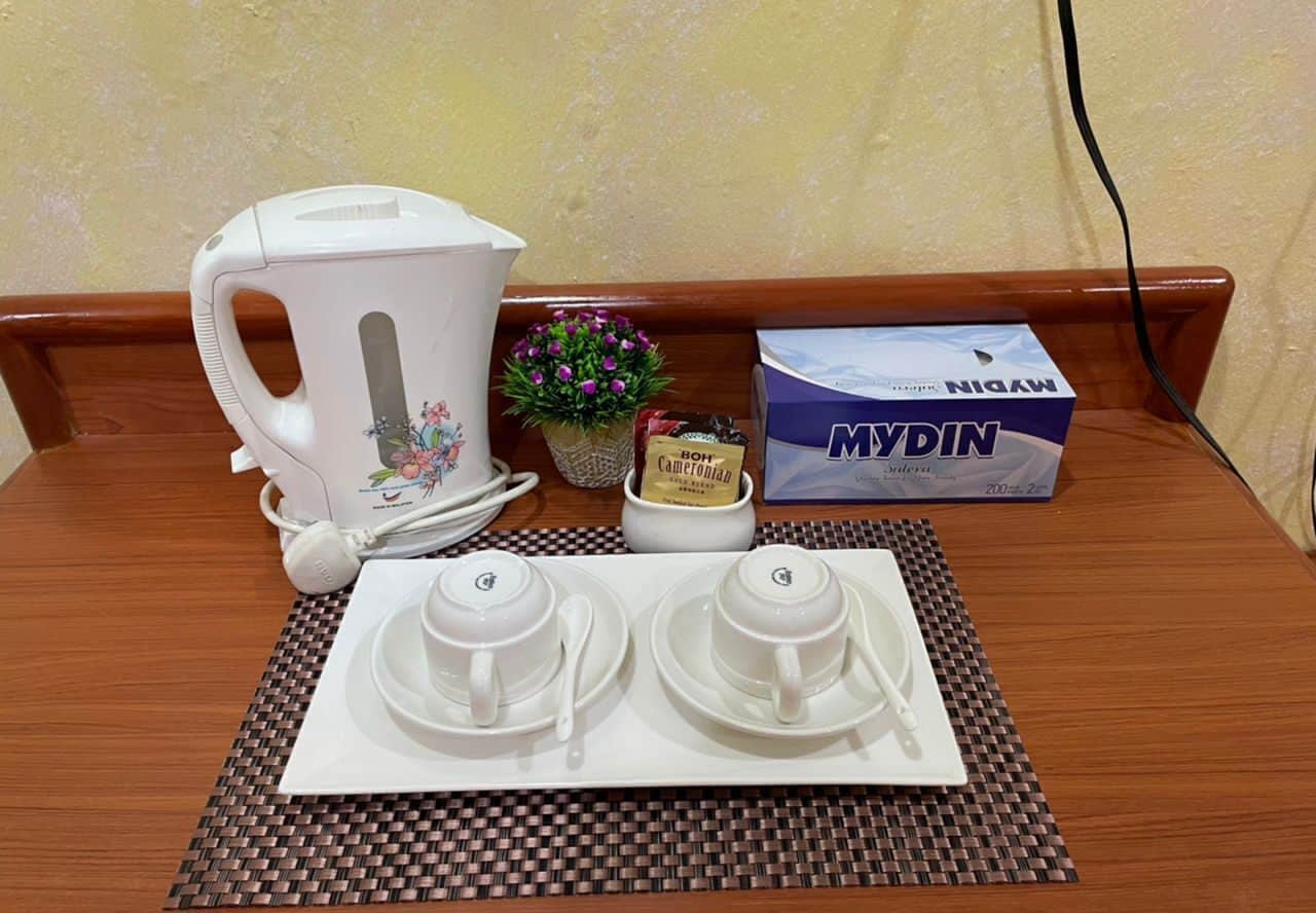 Uma mesa com uma chaleira elétrica branca, uma caixa de café MyDIN, uma pequena planta em vaso e duas xícaras de chá brancas sobre um descanso tecido.