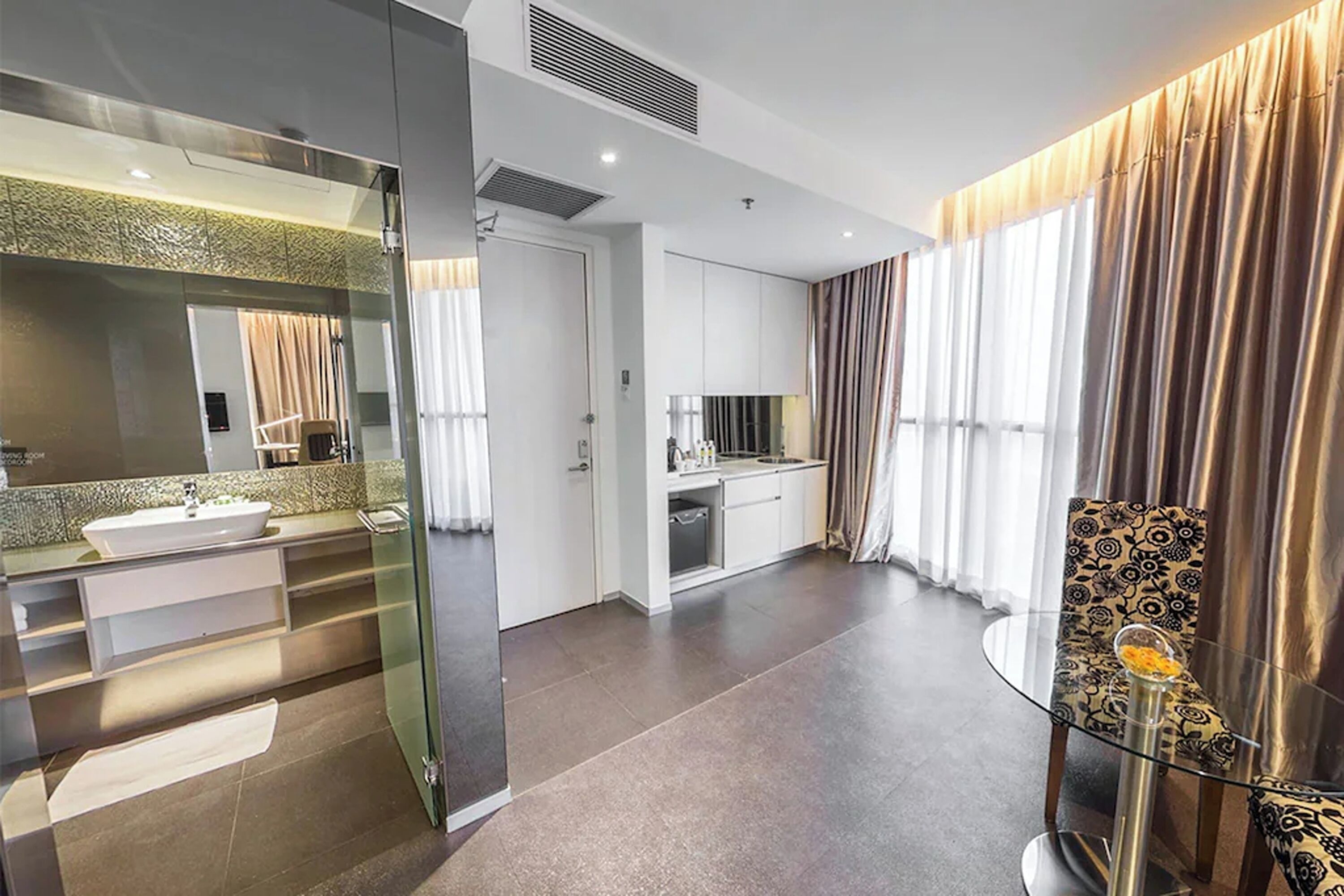 Quarto de hotel moderno com chuveiro de vidro, toucador com espelho grande, pequena cozinha e área de estar com mesa de vidro e cadeira estampada.