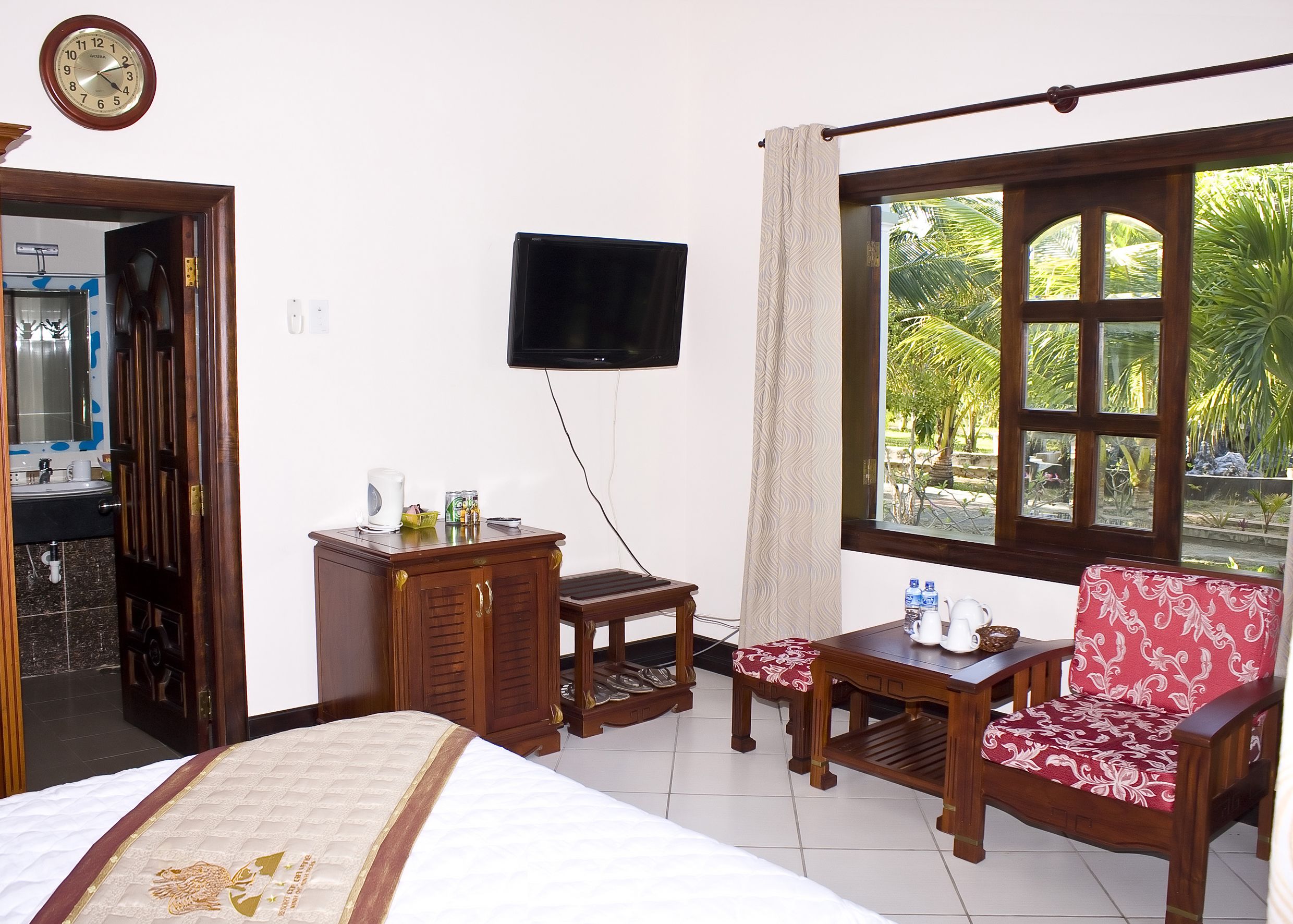 Quarto de hotel com uma cama, penteadeira de madeira, televisão, janela com vista para o verde e uma cadeira vermelha com padrão.