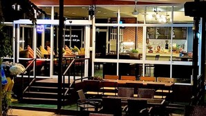 Uma área externa com mesas e cadeiras, uma escadaria que leva a um andar superior e vista do interior de um restaurante, incluindo um balcão e assentos.