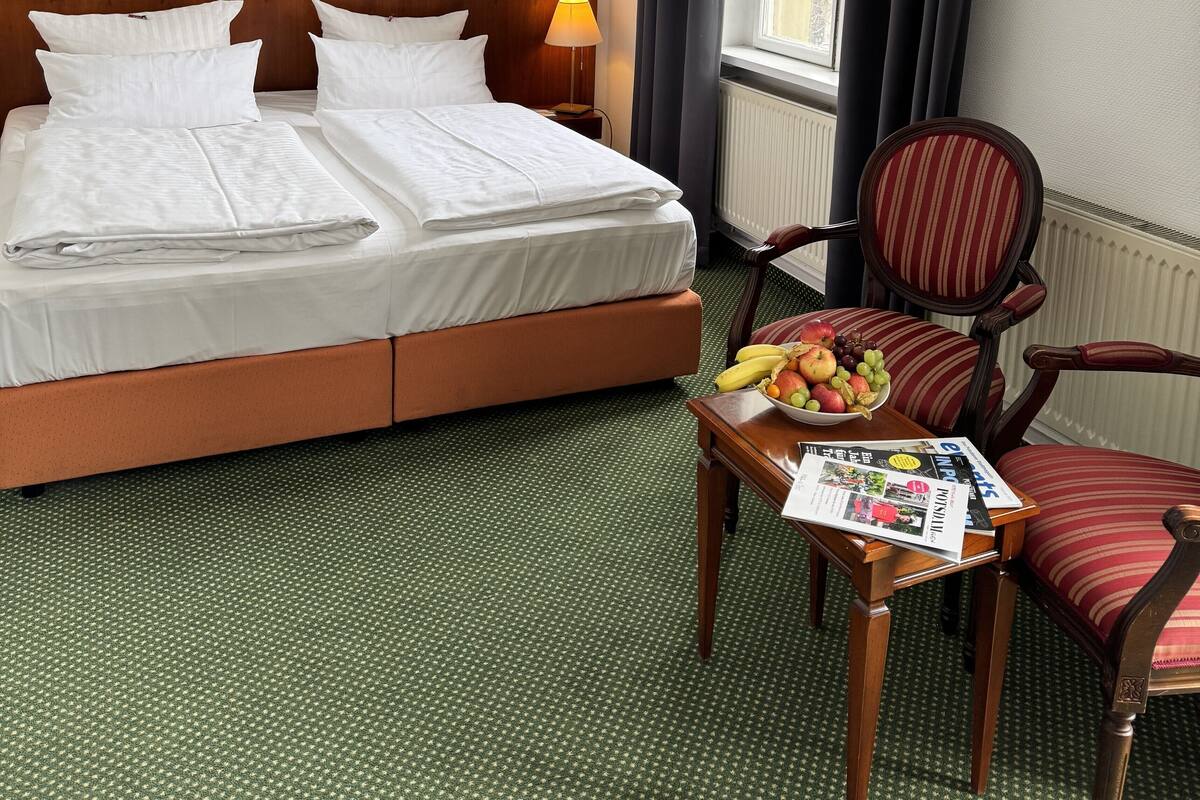 Quarto de hotel com cama bem arrumada, uma mesa de jantar com uma cesta de frutas e uma cadeira.