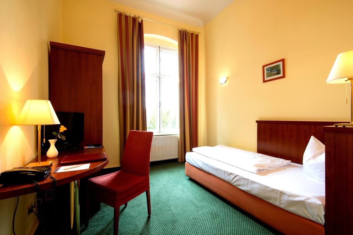 Quarto de hotel com uma cama, uma escrivaninha com luminária, uma cadeira e uma televisão.
