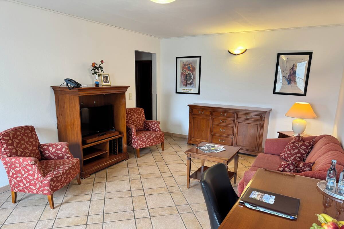 Uma sala de estar com televisão, dois poltronas estampadas, um sofá, um aparador de madeira e uma mesa de jantar com luminária e tigela de frutas.