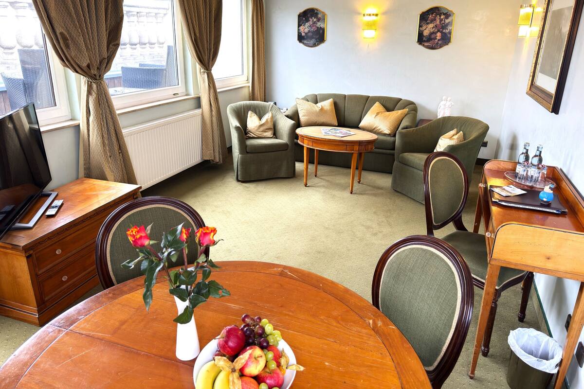 Sala de estar com sofá, poltronas, mesa de centro e mesa de jantar com frutas. Há duas janelas com cortinas, um radiador e luminárias na parede.