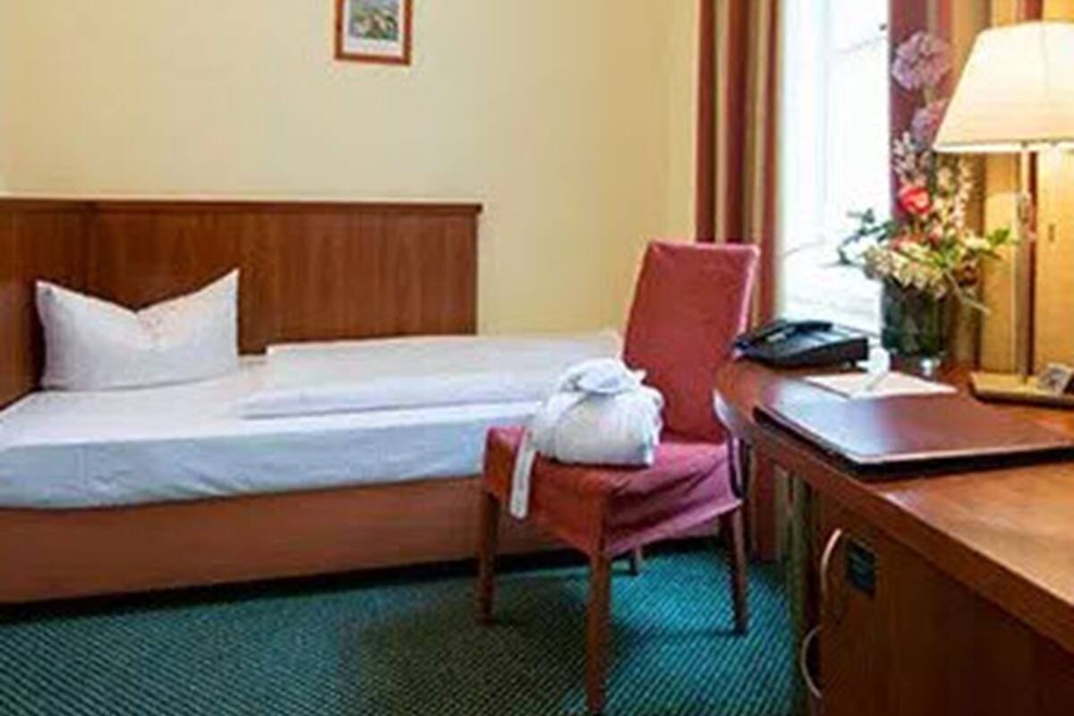 Quarto de hotel com uma cama, escrivaninha, cadeira, luminária e um quadro na parede.