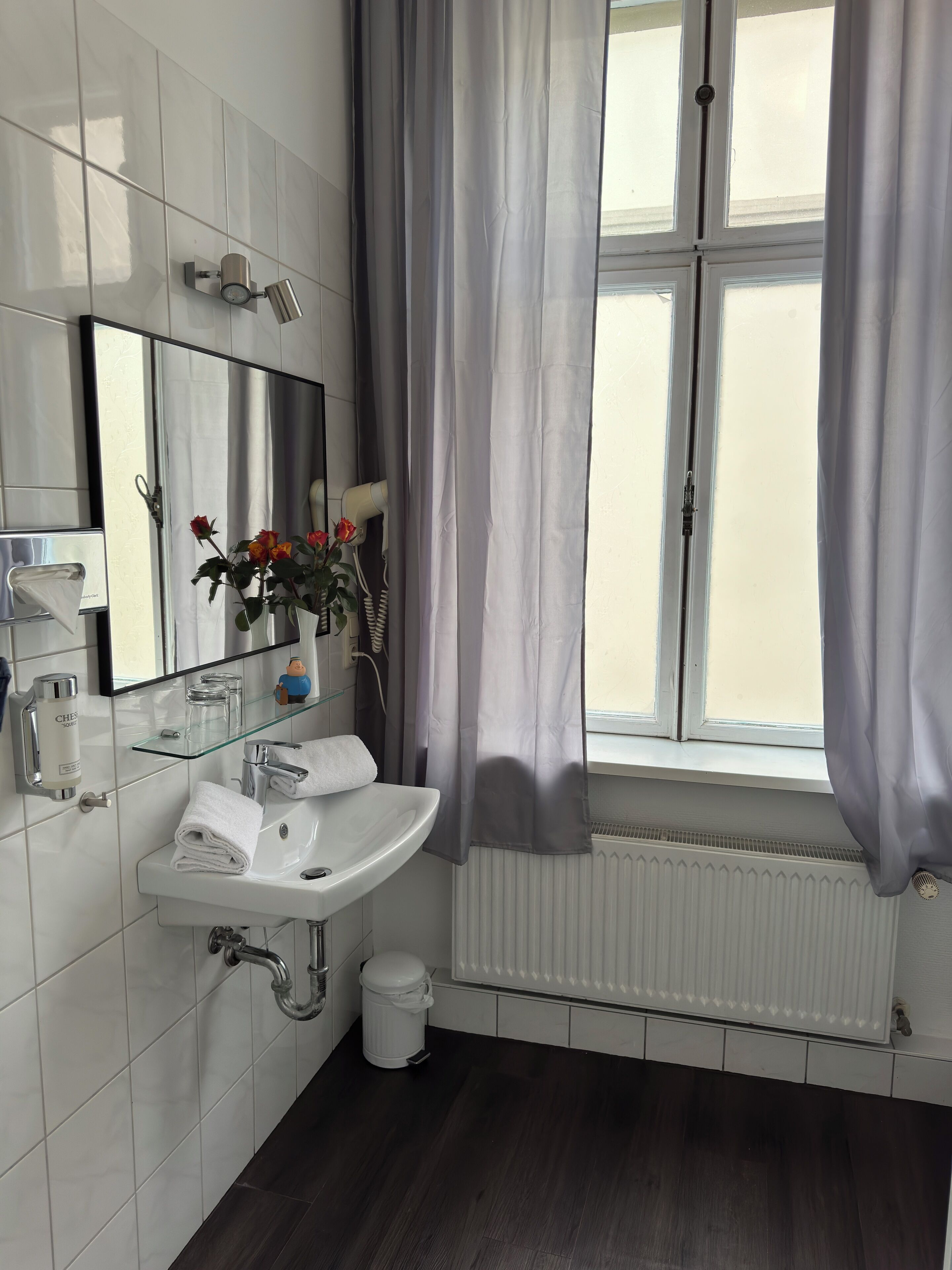 Uma casa de banho moderna com uma sanita branca, espelho e barra de toalhas. Tem uma janela com cortinas, radiador e um cesto de lixo.
