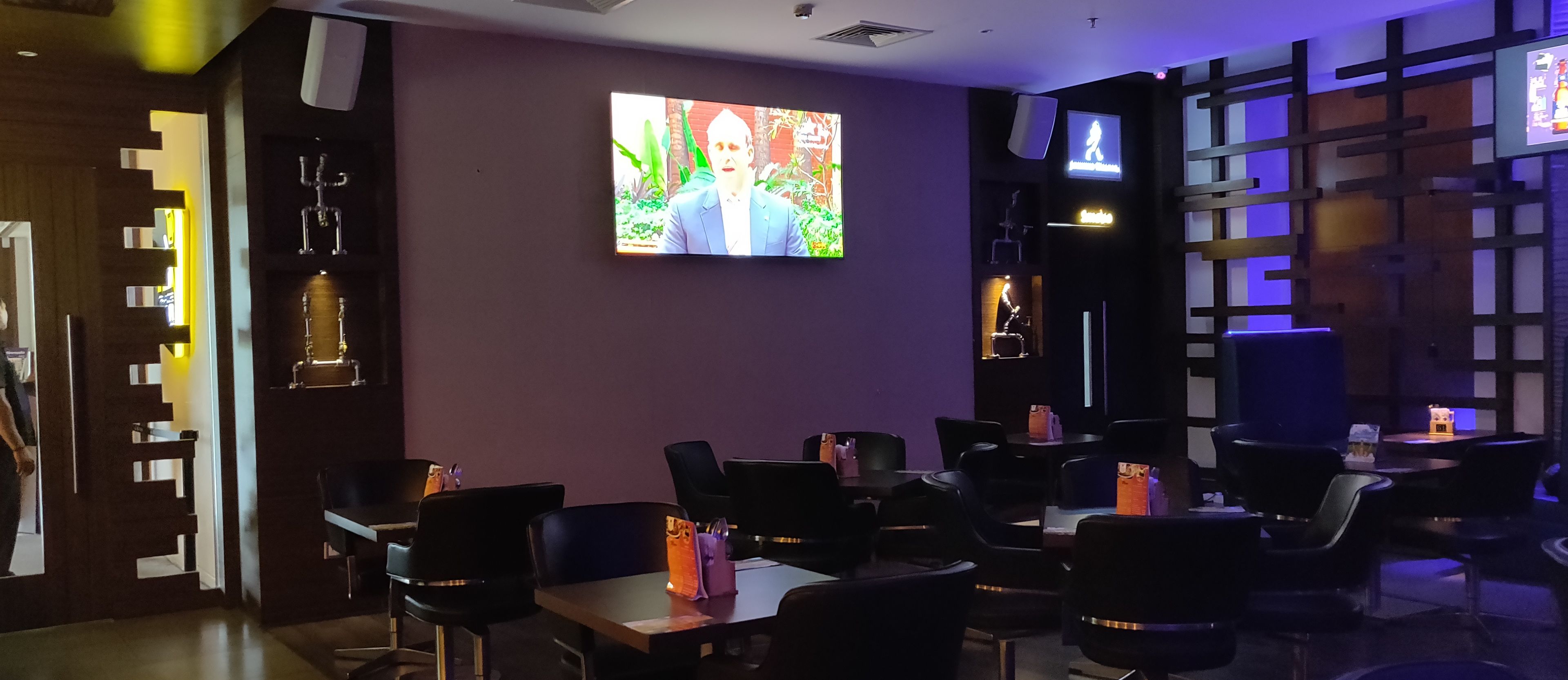 Restaurante com iluminação baixa, mesas e cadeiras, uma tela de televisão exibindo um programa e uma parede decorativa com padrão geométrico.