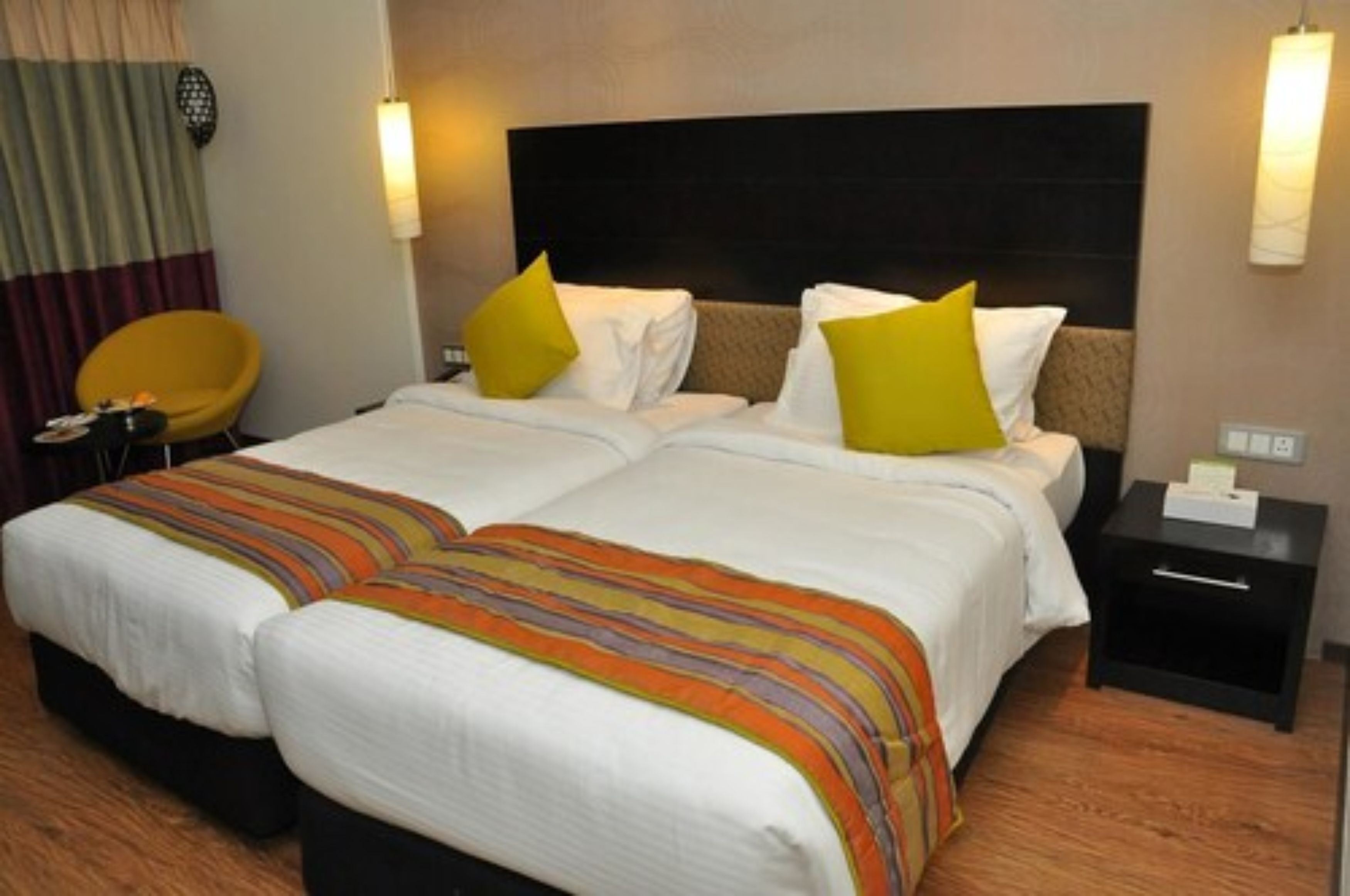 Quarto de hotel com duas camas, uma cadeira, uma mesinha de cabeceira e um abajur.