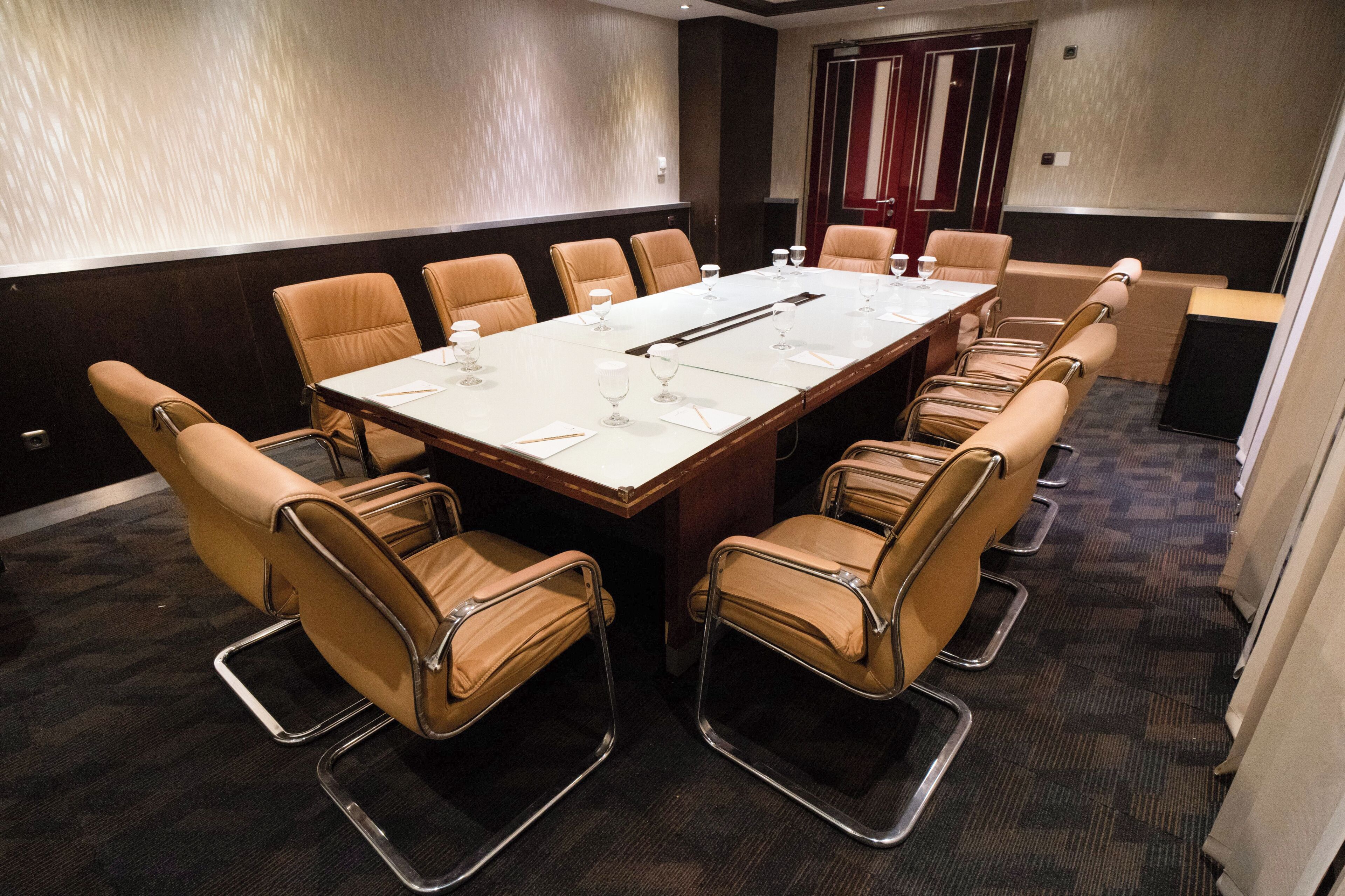 Uma sala de conferências com uma mesa comprida, várias cadeiras e uma porta ao fundo.