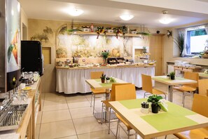 Restaurante com buffet, várias mesas, cadeiras e área de cozinha com um mural decorativo.