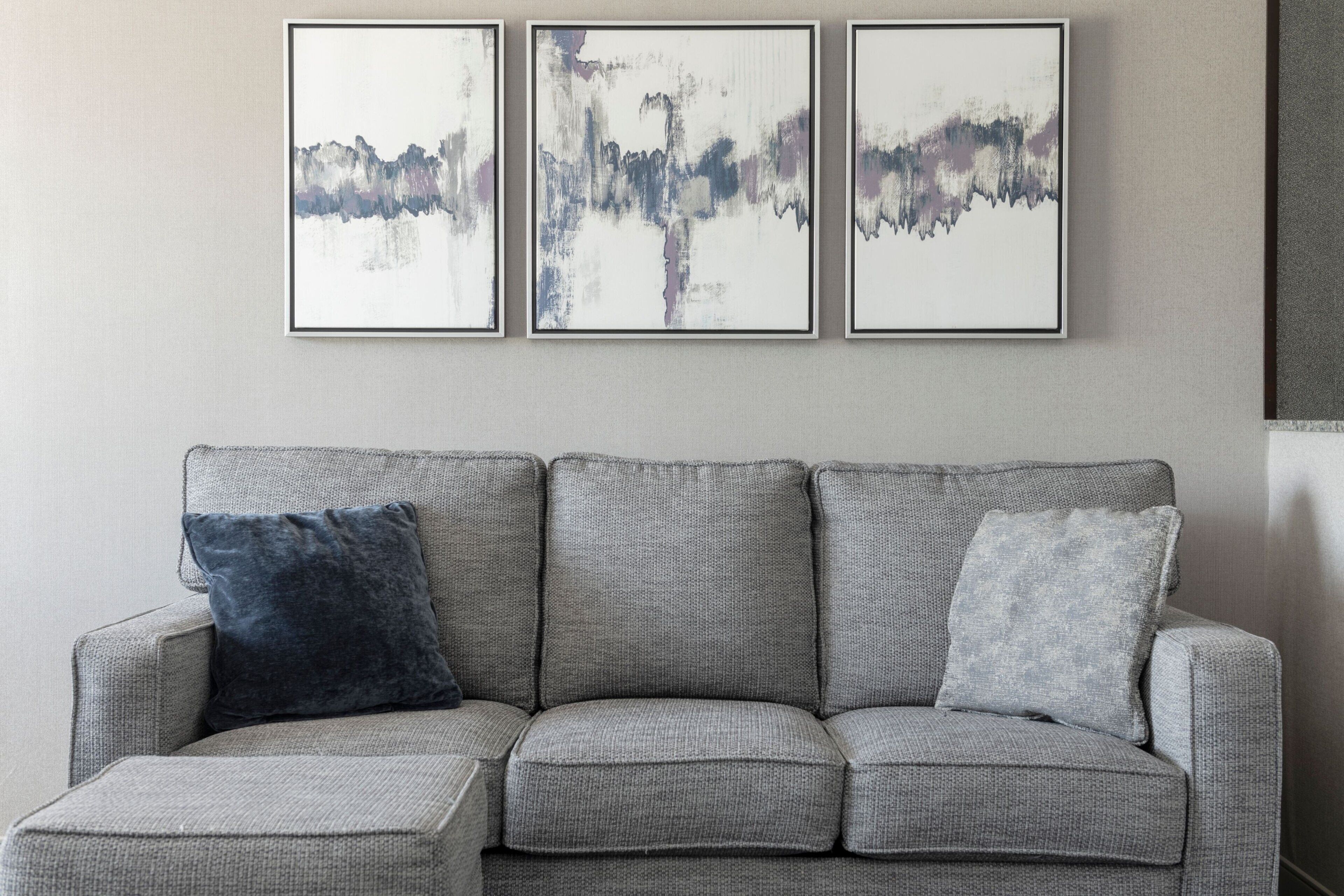 Um sofá cinzento com duas almofadas, um quadro abstrato emoldurado acima e uma parede lisa.