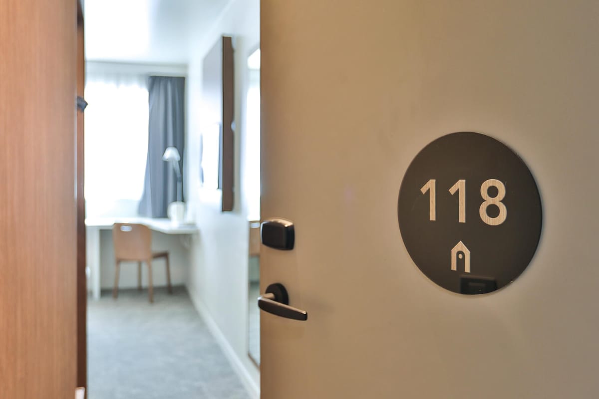 Porta de quarto de hotel com placa indicando o quarto 118, maçaneta e vista parcial de um quarto com uma escrivaninha e cadeira.