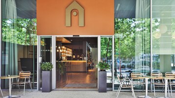 Um restaurante com entrada aberta, mesas internas e um bar visível através das portas de vidro.