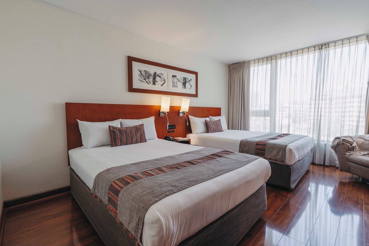 Quarto de hotel com duas camas, cabeceira de madeira, quadro emoldurado acima das camas e uma janela grande com cortinas finas.