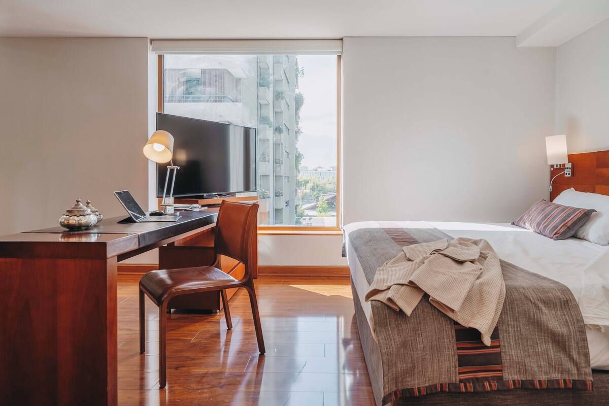 Quarto de hotel com cama, escrivaninha com laptop, cadeira e uma janela grande com vista para a cidade.