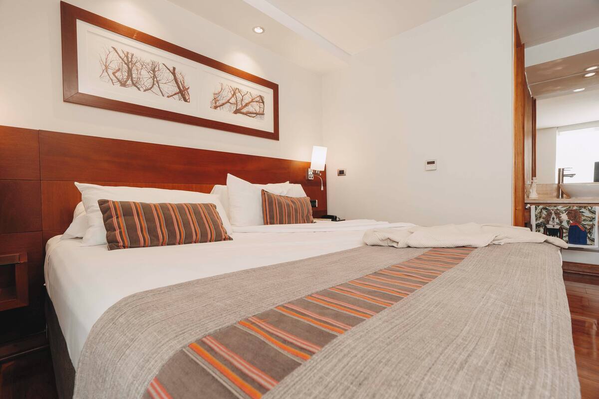 Quarto de hotel com uma cama grande, cabeceira de madeira, uma obra de arte emoldurada acima da cama e uma mesa de cabeceira com uma luminária.