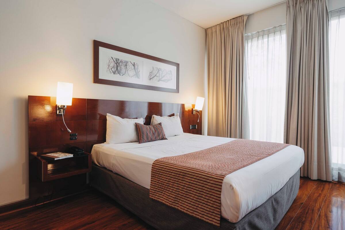 Quarto de hotel com uma cama grande, duas mesas de cabeceira, um criado-mudo, uma obra de arte emoldurada acima da cama e uma janela com cortinas.