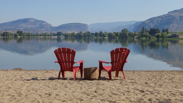 Duas cadeiras Adirondack vermelhas numa praia arenosa, com um lago calmo e montanhas ao fundo.