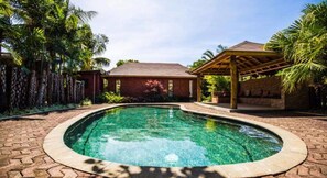 Piscina com borda de tijolos, cercada por palmeiras e uma pérgula de madeira.