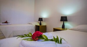 Quarto de hotel com duas camas, cada uma com um cobertor branco e uma luminária em uma mesa de cabeceira de madeira.