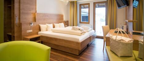 Quarto de hotel moderno com uma cama grande, uma escrivaninha com cadeira, uma poltrona verde e uma TV fixada na parede.