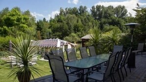 Uma mesa com tampo de vidro e cadeiras, cercada por um jardim com árvores e um gazebo ao fundo.