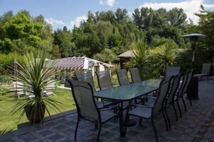 Uma mesa com tampo de vidro e cadeiras, cercada por um jardim com árvores e um gazebo ao fundo.