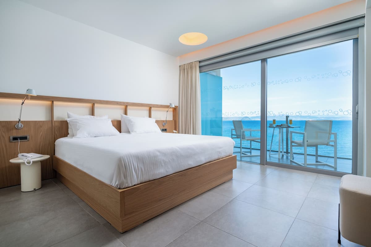 Quarto de hotel moderno com uma cama grande, uma mesinha de cabeceira com luminária, uma pequena mesa com cadeiras e uma varanda com vista para o mar.