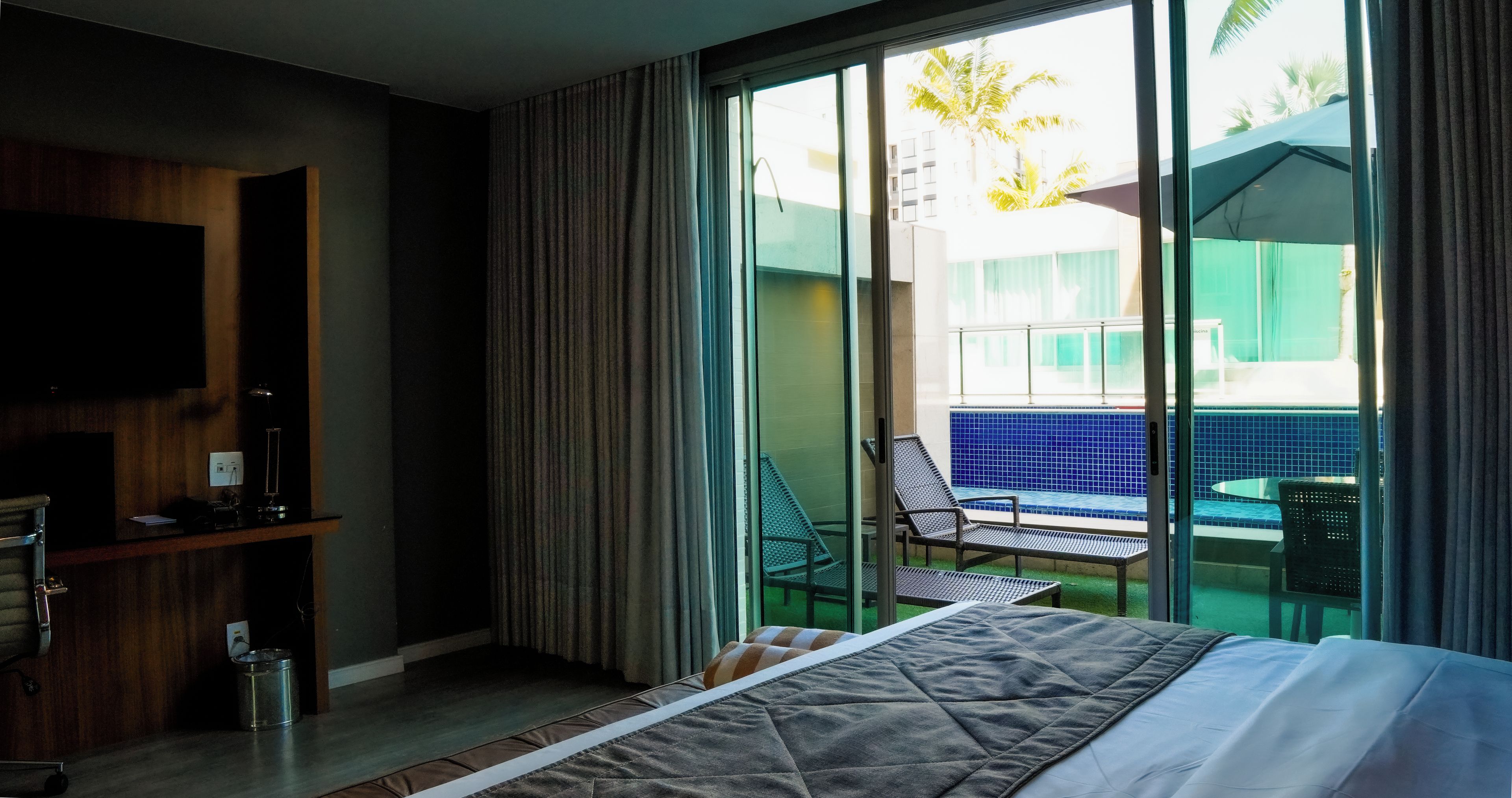 Quarto de hotel com cama, escrivaninha, TV e vista para a piscina e palmeiras.