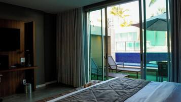 Quarto de hotel com cama, escrivaninha, TV e vista para a piscina e palmeiras.