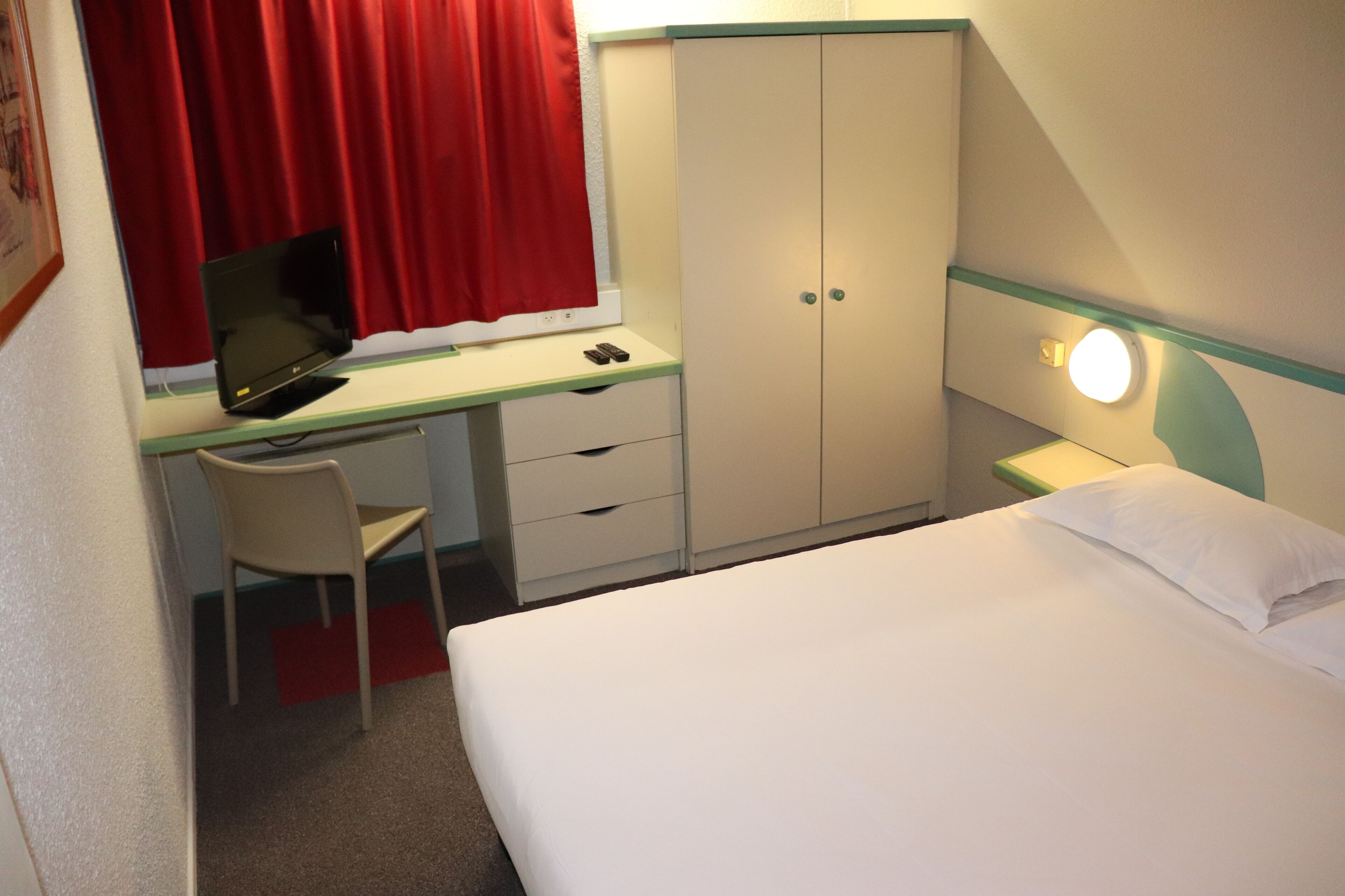 Quarto de hotel com cama, escrivaninha, computador, cadeira, guarda-roupa e abajur.