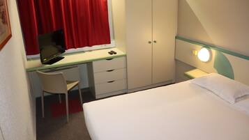 Quarto de hotel com cama, escrivaninha, computador, cadeira, guarda-roupa e abajur.