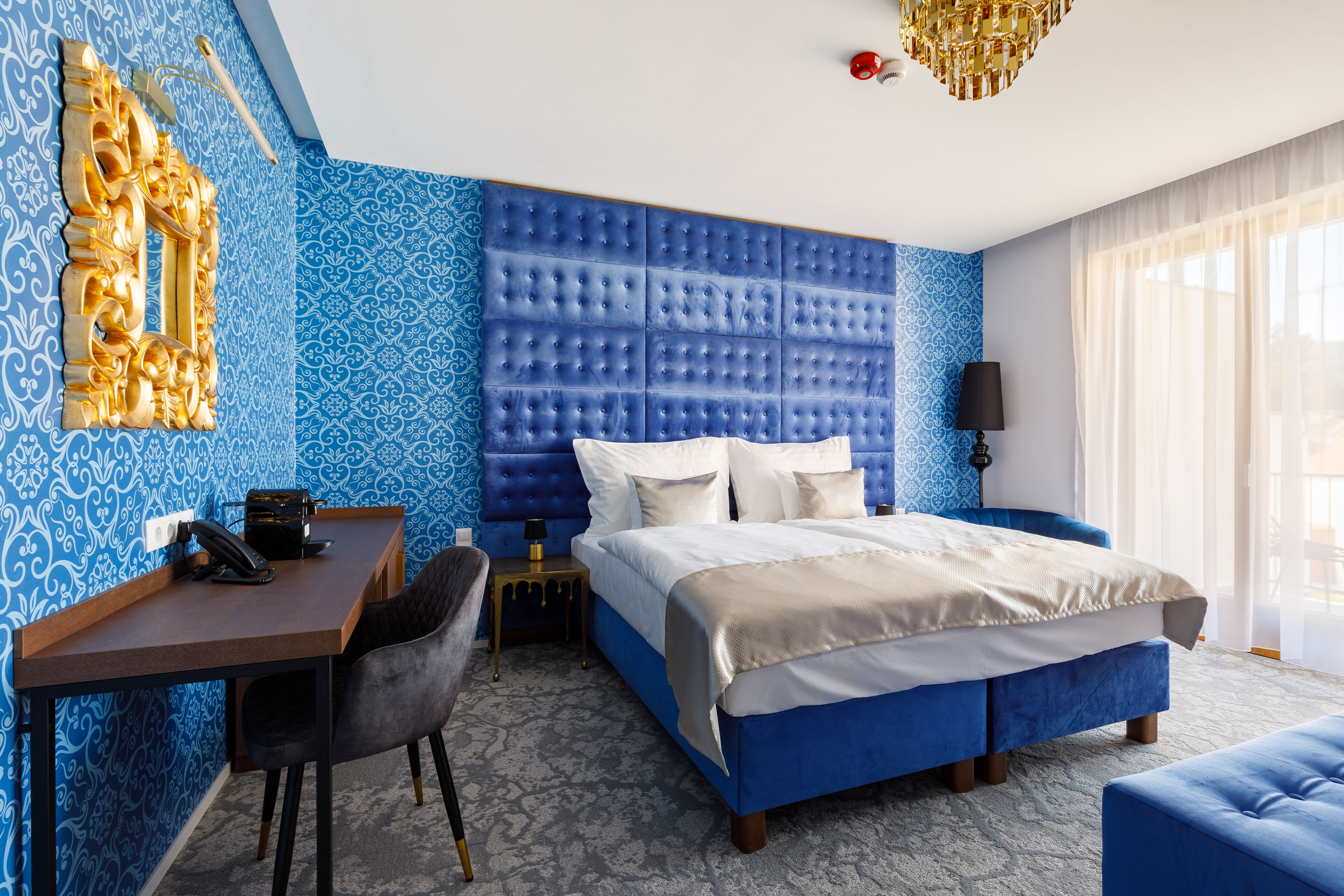 Quarto de hotel com parede estampada em azul, cabeceira estofada, escrivaninha com telefone, uma cadeira e cama com roupa de cama branca.