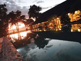 Uma área de piscina ao ar livre tranquila com vista para o pôr do sol, cercada por árvores e um edifício com janelas iluminadas.