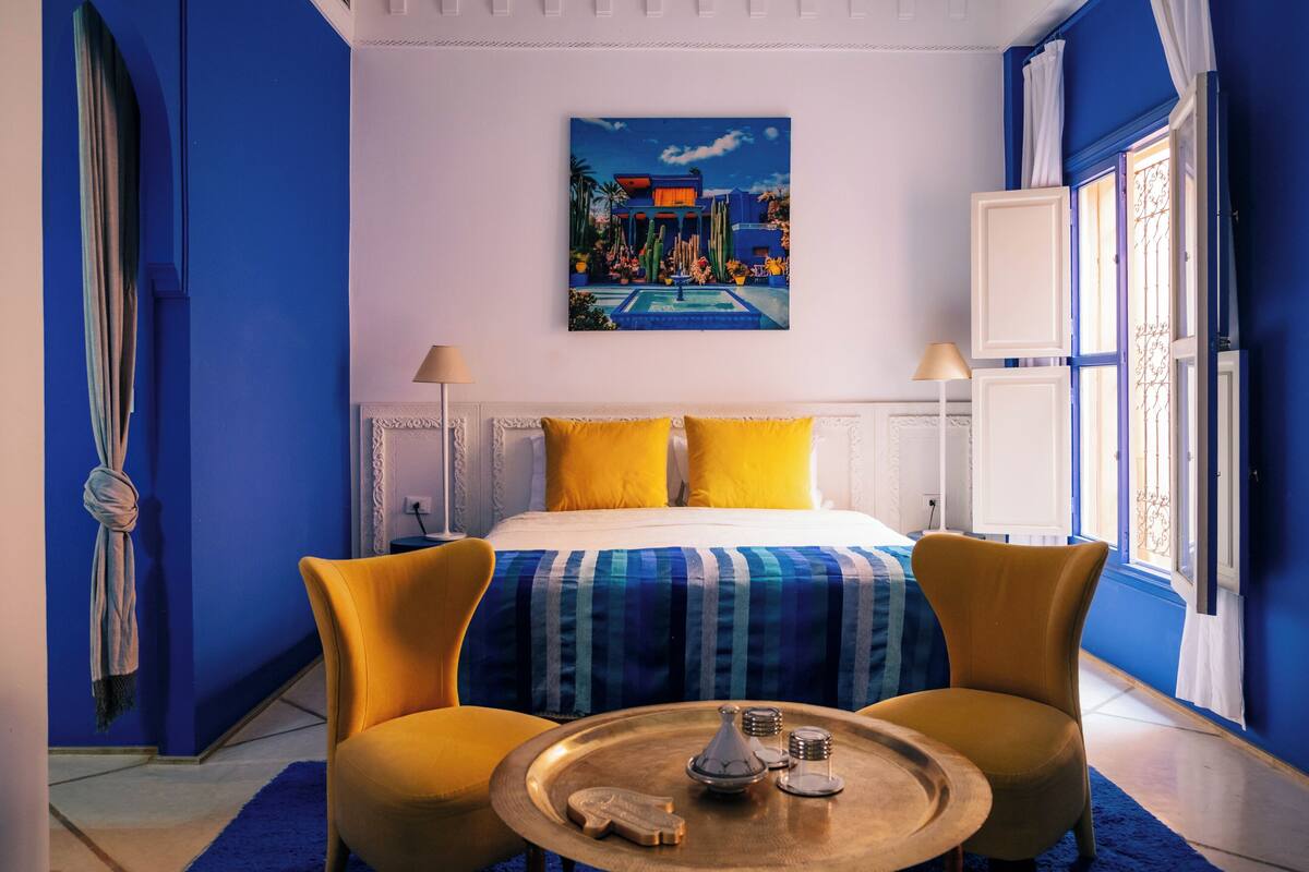 Quarto de hotel com uma cama, duas poltronas amarelas, uma mesa redonda com um tabuleiro e um quadro de uma piscina na parede.