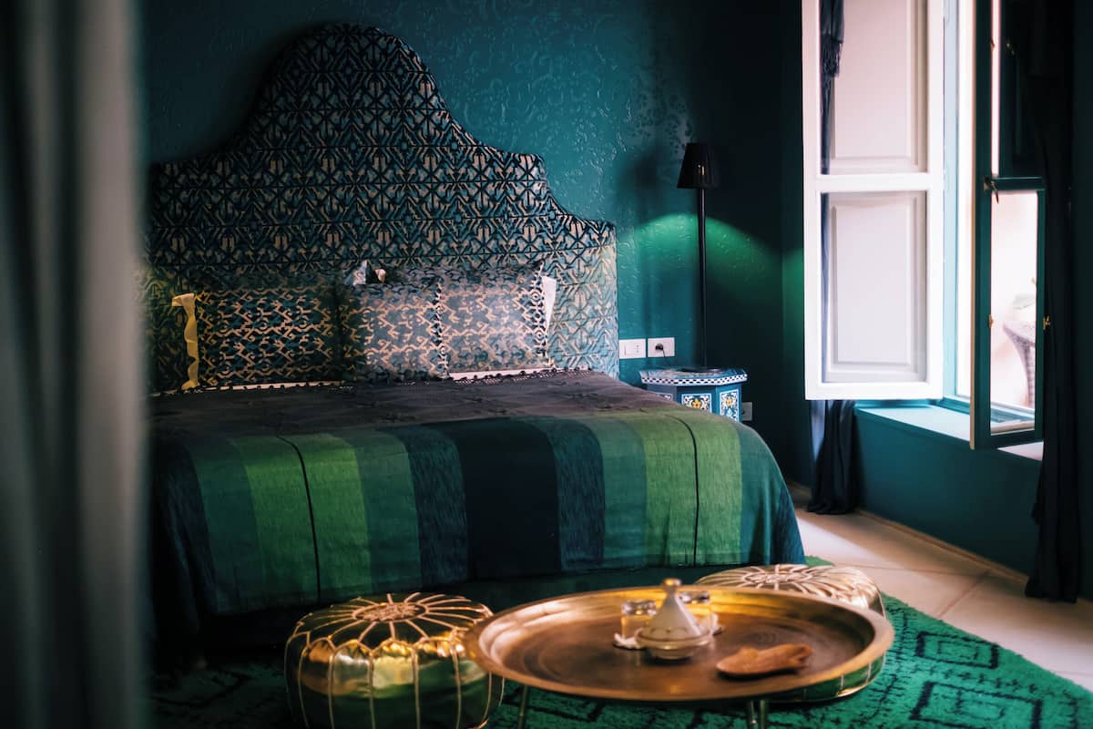Quarto com cabeceira estampada, colcha de cama listrada em verde, mesa redonda pequena com bandeja e janela com cortinas.