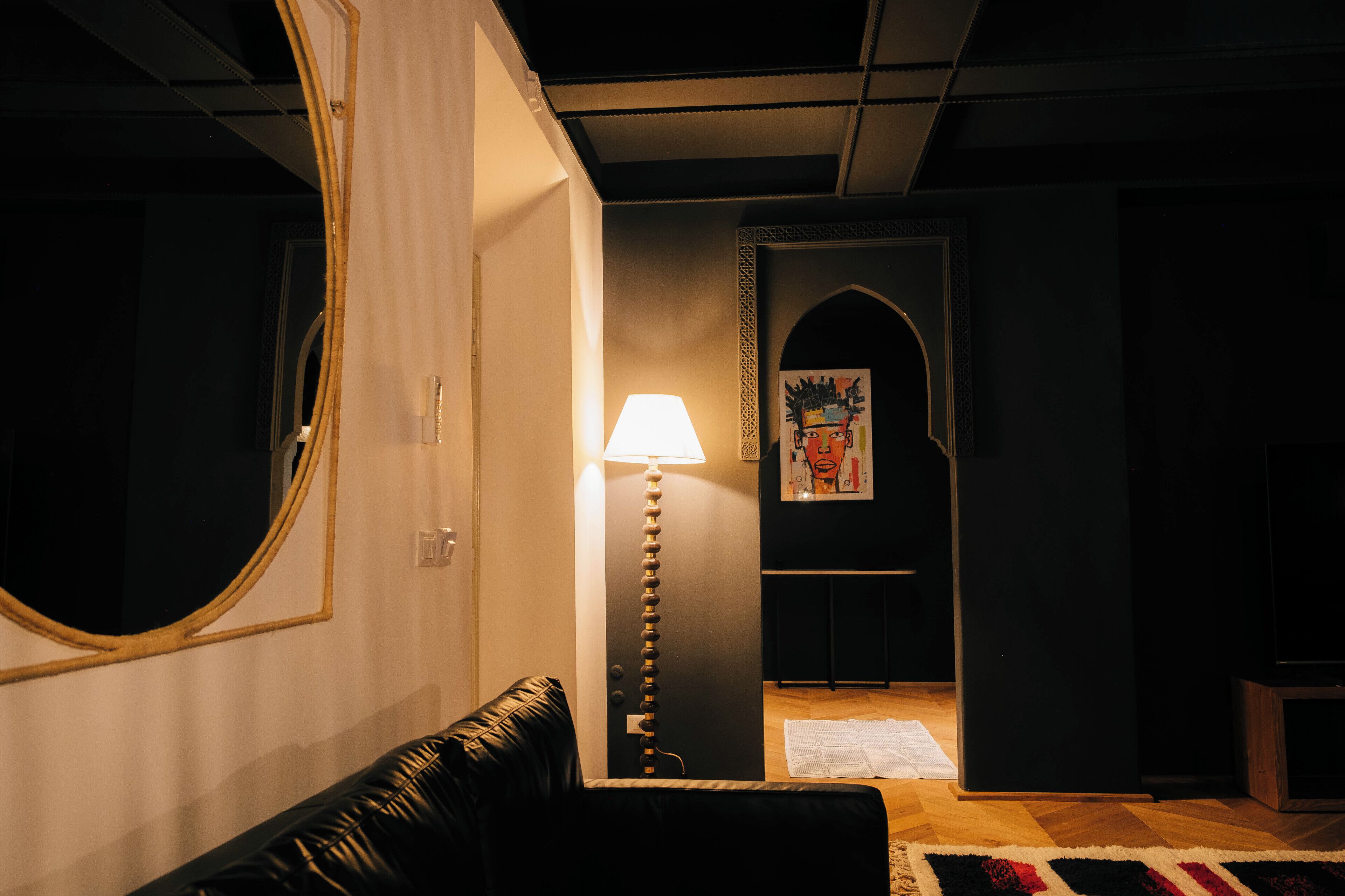 Quarto com sofá de couro preto, luminária, espelho e porta que dá para outro cômodo.