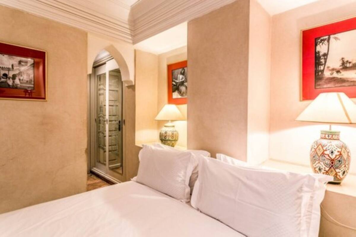 Quarto de hotel com uma cama, duas mesas de cabeceira com abajures, uma porta e quadros nas paredes.