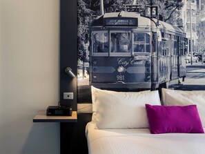 Quarto de hotel com cama, almofadas, mesa de cabeceira com rádio e um mural na parede com um bonde.