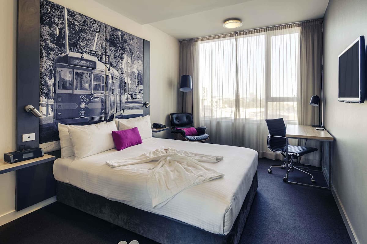 Quarto de hotel com cama, escrivaninha, cadeira, televisão e um pôster grande na parede.
