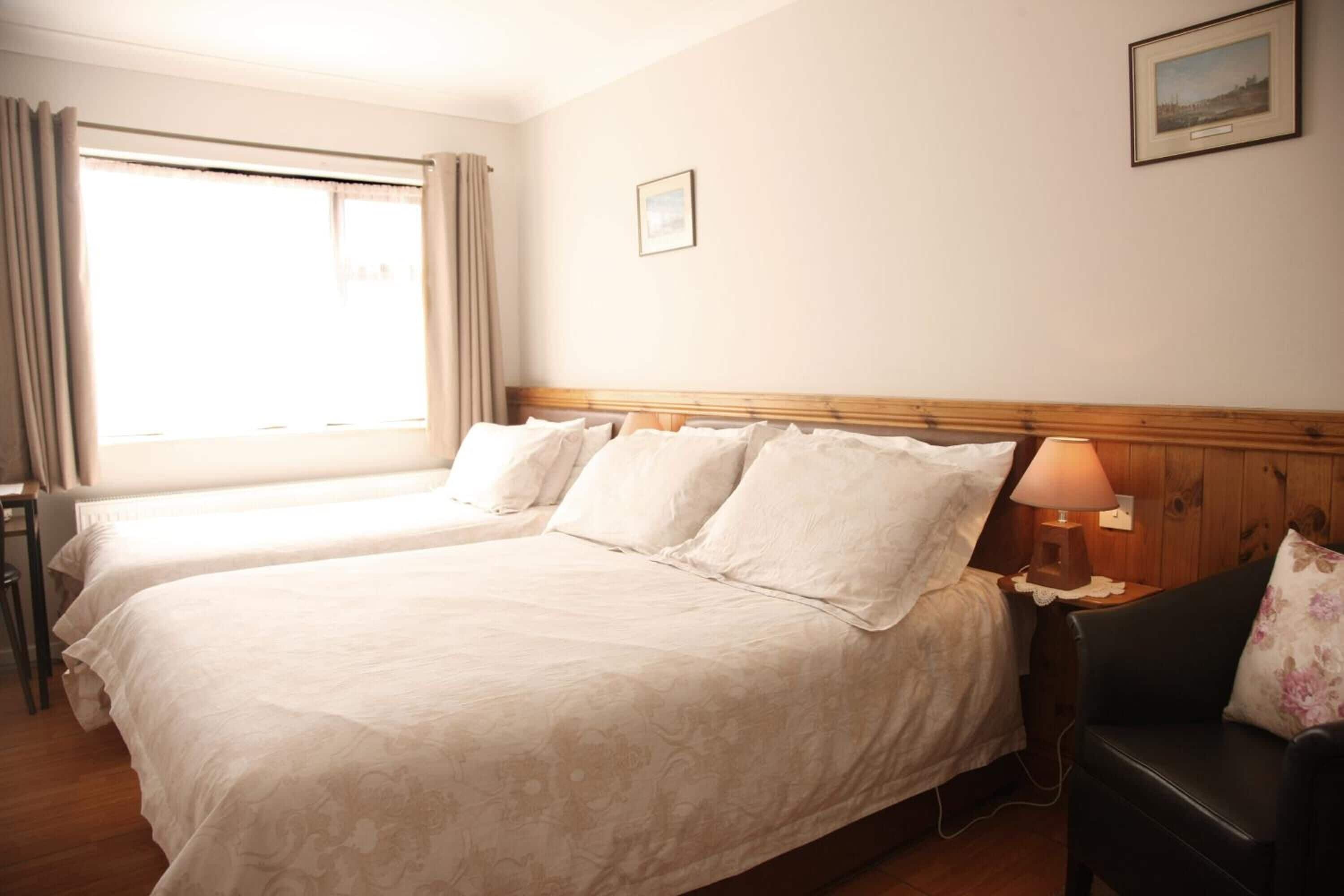 Quarto de hotel com duas camas, cabeceira de madeira, mesa de cabeceira com abajur, quadro na parede e janela com cortinas.