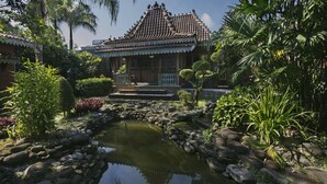 Um edifício tradicional com telhado em tijolos, rodeado por um jardim com lago e diversas plantas.