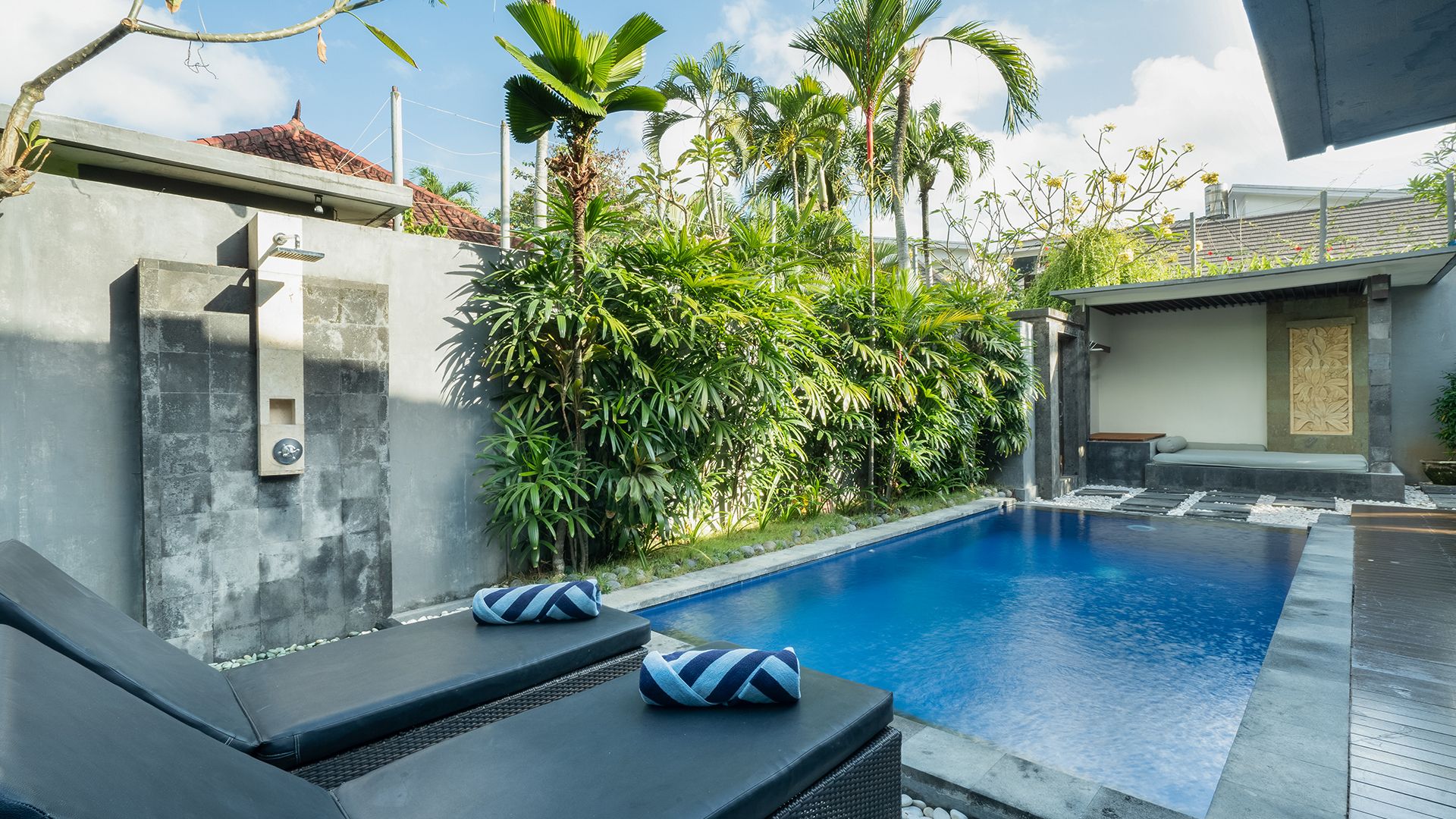 Piscina com muro de pedra e plantas tropicais, área de estar com almofadas e um edifício moderno ao fundo.