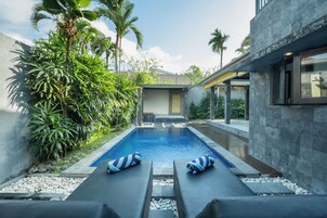 Uma área de piscina moderna com duas espreguiçadeiras e uma piscina pequena, cercada por vegetação exuberante e um muro de pedra.