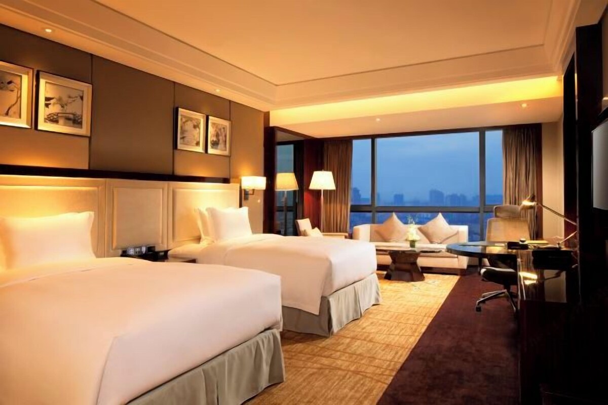 Quarto de hotel com duas camas, uma escrivaninha, uma cadeira e uma janela grande com vista para a cidade.