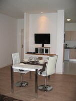Uma área de refeições com uma mesa posta para quatro pessoas, uma televisão de tela plana na parede e uma cozinha ao fundo.